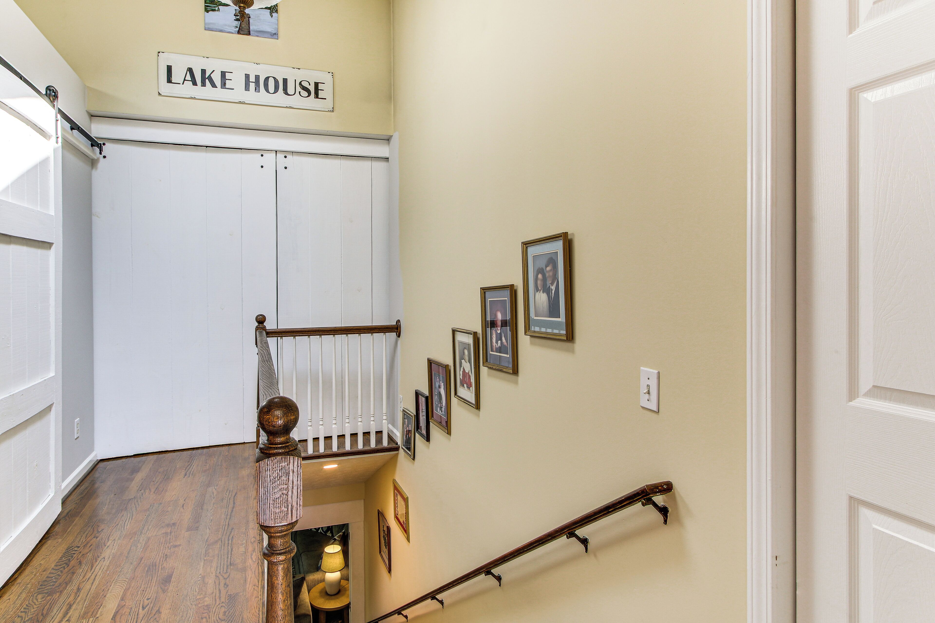 Uma escadaria com corrimão de madeira, uma porta branca com a placa "LAKE HOUSE" e várias fotos emolduradas na parede.