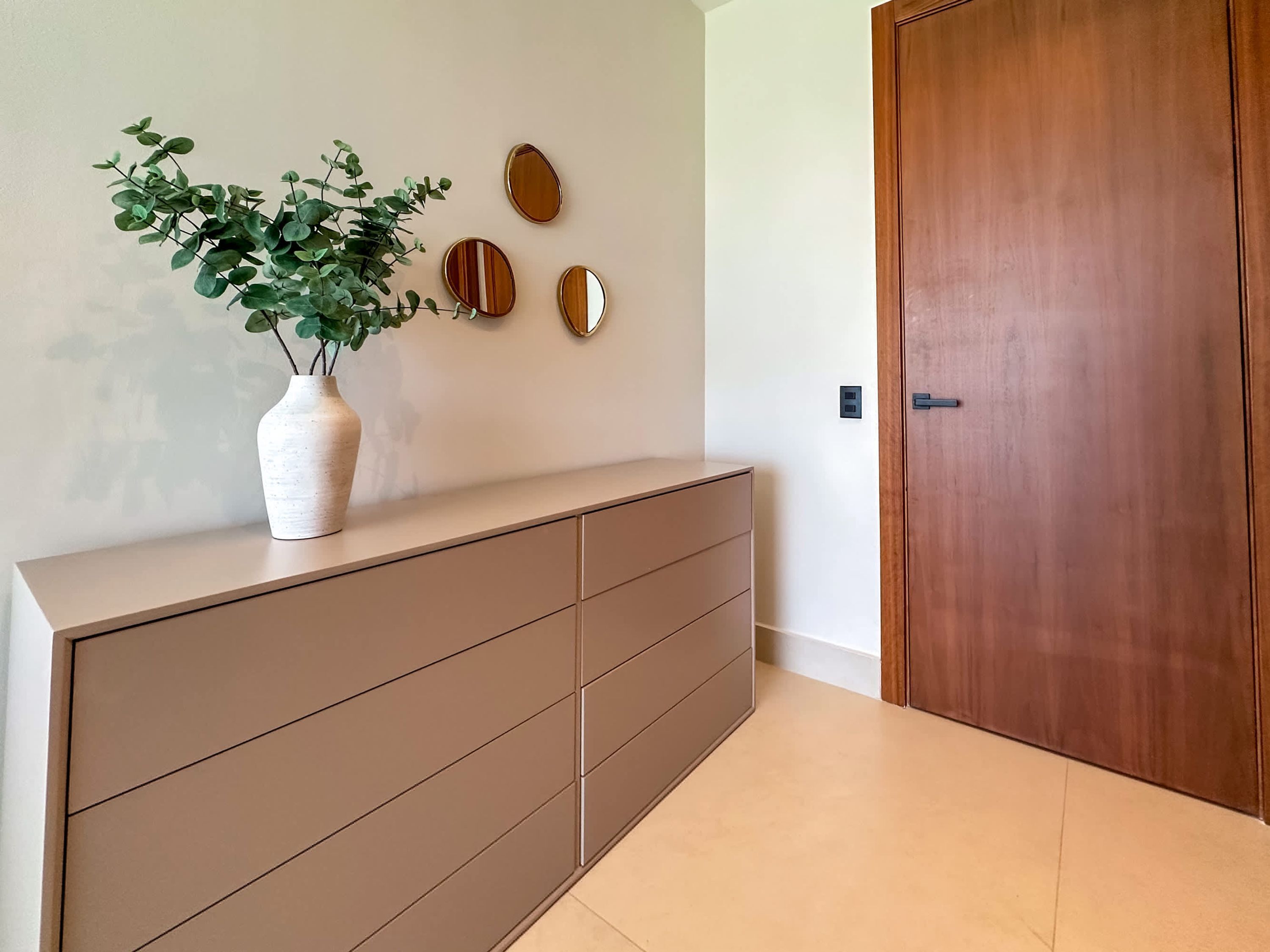 Um corredor com um armário bege, uma planta em vaso e três espelhos decorativos na parede.