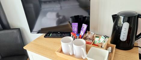 Quarto de hotel com televisão, máquina de café, bandeja com lanches e uma caneca branca.