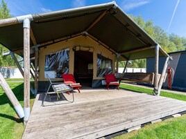 Área externa coberta com duas cadeiras vermelhas e uma mesa, cercada por um deck de madeira e gramado.