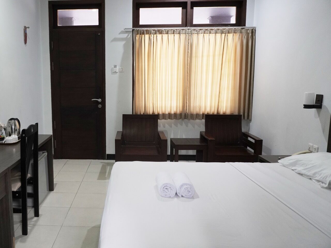 Quarto de hotel com cama, duas cadeiras, uma mesa pequena, escrivaninha com chaleira e janela com cortinas.