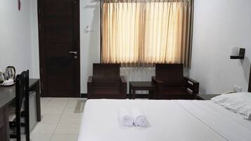 Quarto de hotel com cama, duas cadeiras, uma mesa pequena, escrivaninha com chaleira e janela com cortinas.