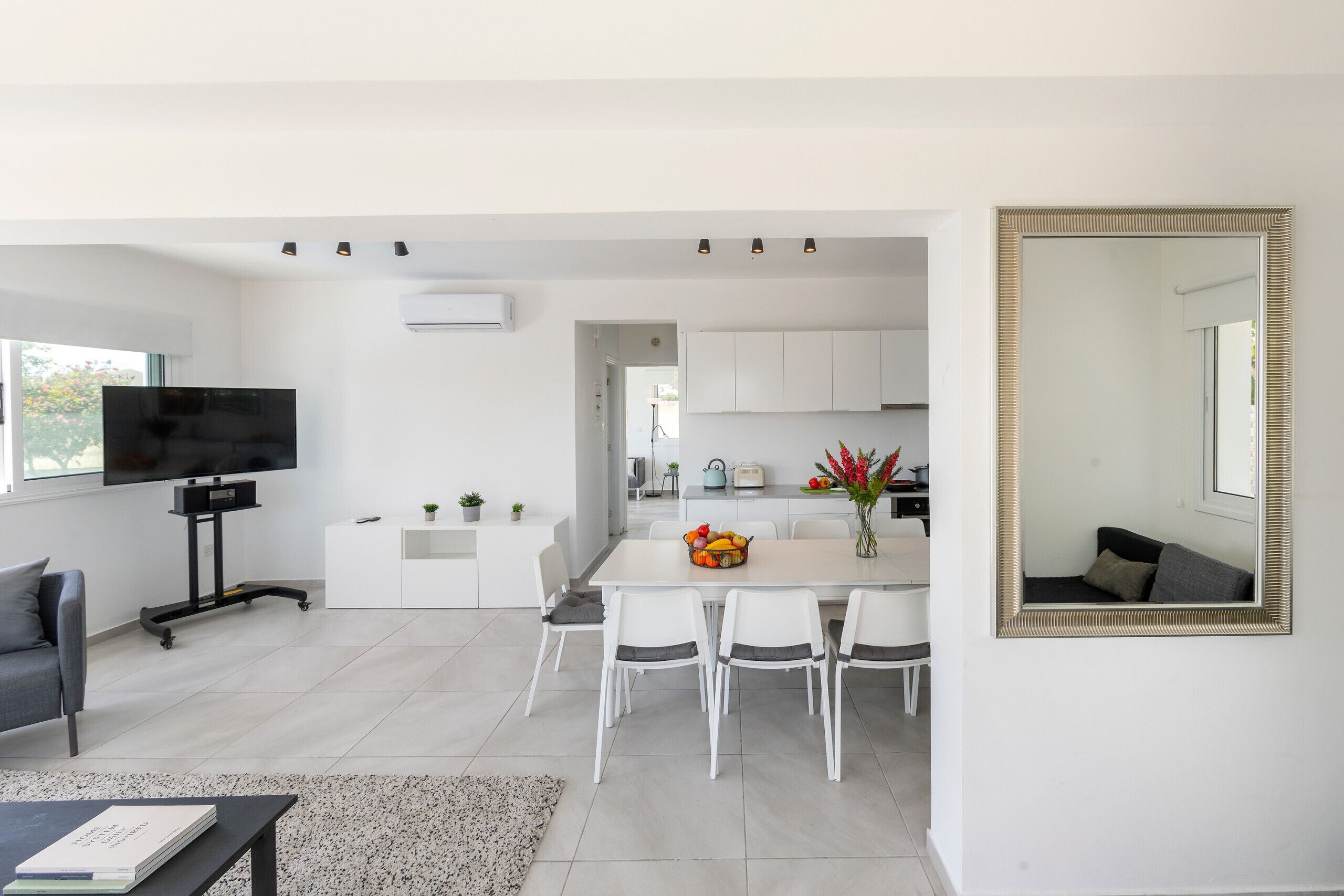 Uma sala de estar moderna com uma TV de tela plana, uma área de jantar com mesa e cadeiras, e uma cozinha com armários e bancada.
