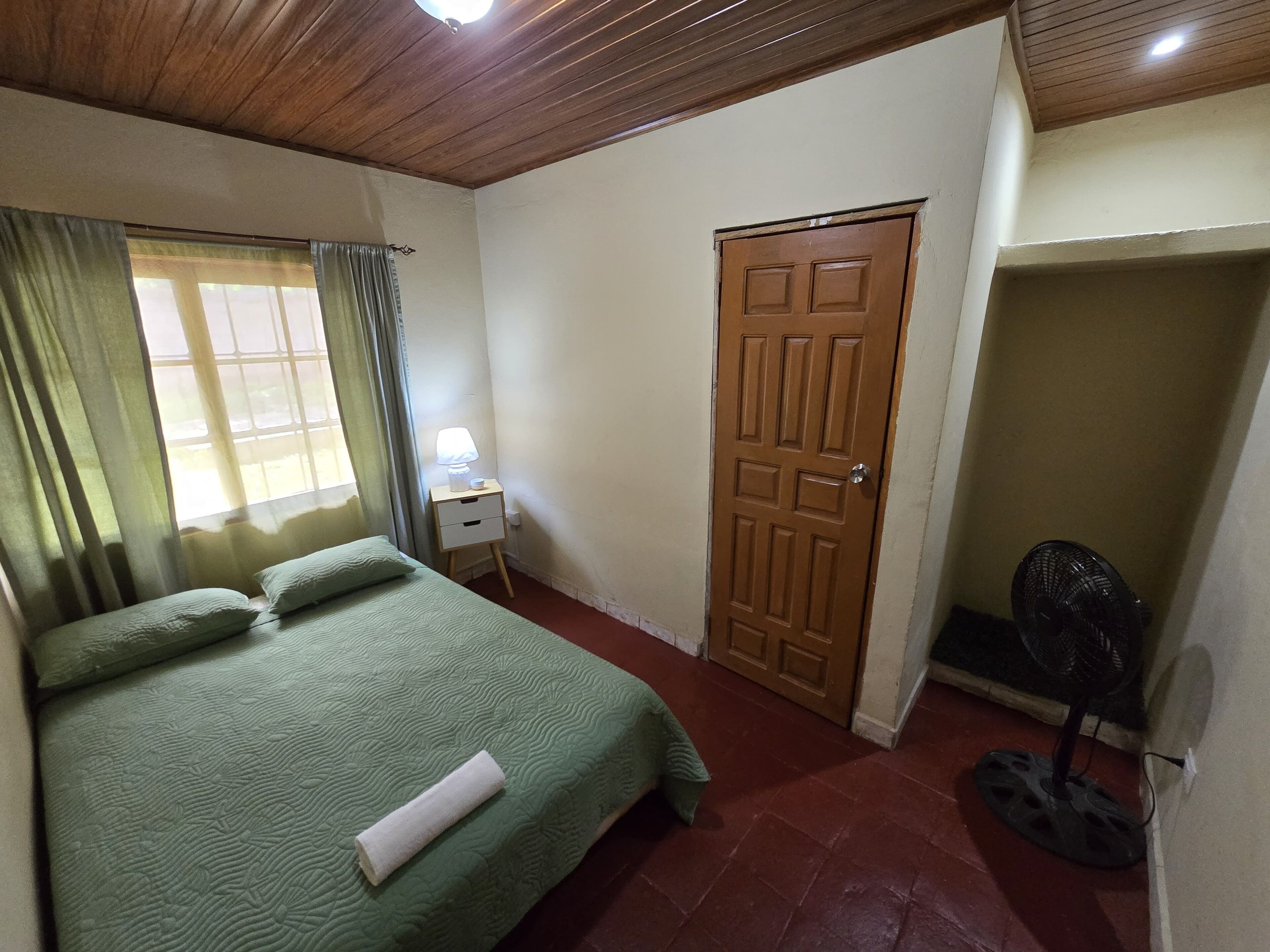 Um quarto com uma cama, mesa de cabeceira, porta e ventilador.