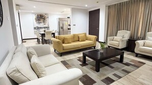 Uma sala de estar moderna com sofá, poltronas e uma mesa de centro. Ao fundo, há uma área de jantar com mesa e cadeiras.