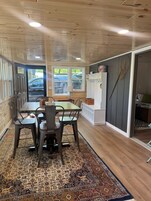 Interior acolhedor de uma cabana com teto de madeira, uma mesa de jantar com cadeiras, um tapete estampado e uma grande janela com vista para o exterior.