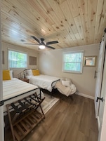 Quarto compacto com duas camas, ventoinha no teto e duas janelas com persianas.