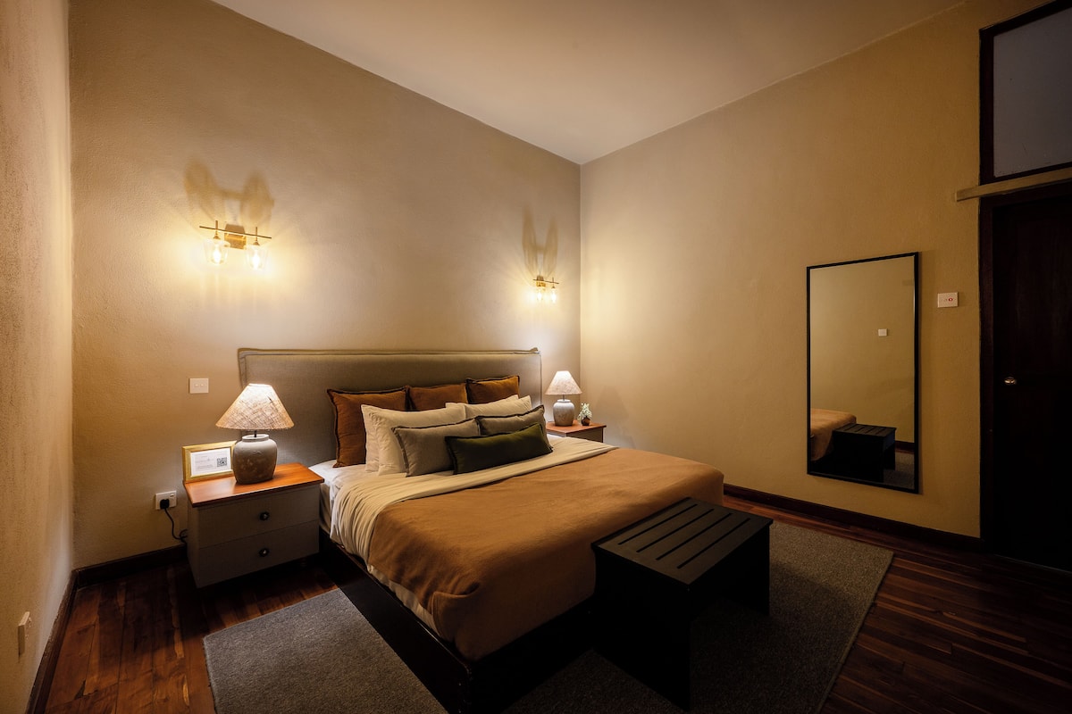Quarto com cama, mesas de cabeceira, luminárias, espelho e um banco de madeira.