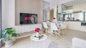Uma sala moderna com televisão de tela plana, área de jantar com cadeiras brancas, cozinha com armários de madeira e uma planta em vaso.