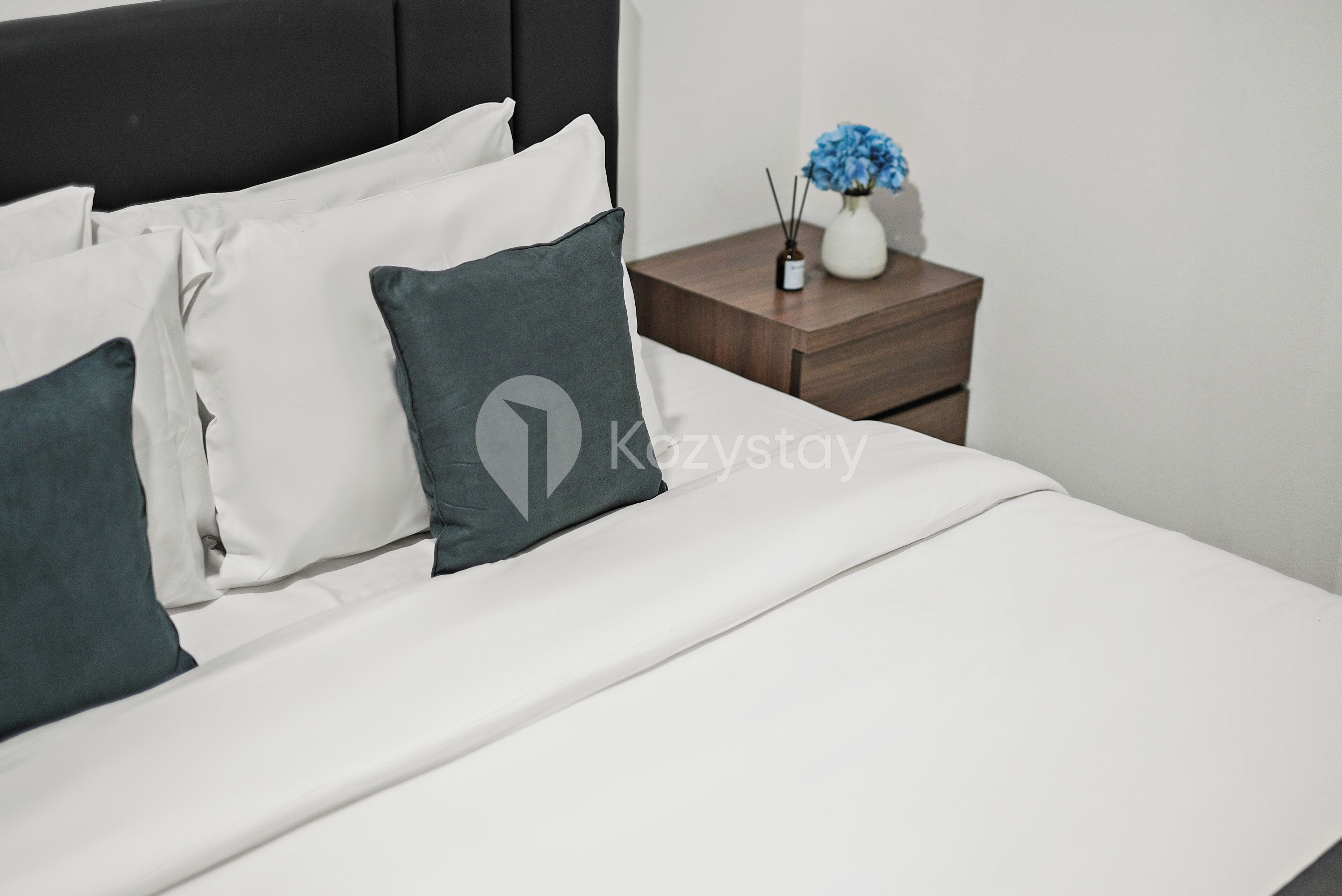 Cama bem arrumada com almofadas brancas e verde-escuras, mesa de cabeceira de madeira com um vaso de flores azuis e um pequeno frasco de incenso.