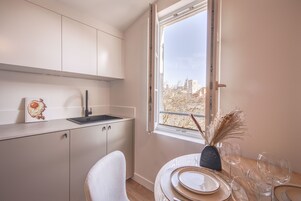 Uma cozinha moderna com armários brancos, uma janela com vista para edifícios e um espaço de refeições com um vaso de plantas secas.