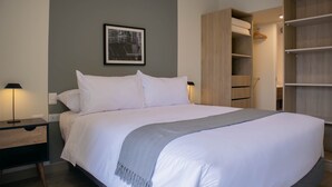Cama bem arrumada com lençóis brancos e manta cinza, mesa de cabeceira com abajur, quadro na parede e guarda-roupa de madeira com espaço para pendurar roupas.