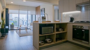 Cozinha moderna equipada com micro-ondas, forno e máquina de café. Possui área de jantar com mesa e cadeiras, além de uma grande janela com vista para a cidade.