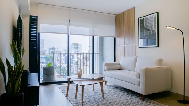 Uma sala de estar moderna com um sofá branco, uma mesa pequena de madeira e um abajur. Há uma janela grande com vista para a paisagem urbana.