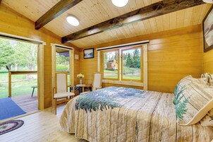 Quarto de cabana de madeira com cama, cadeira, mesa pequena e janela com vista para um quintal.