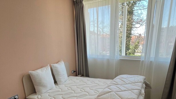 Quarto com cama, mesa de cabeceira, cadeira e janela com cortinas.