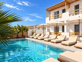 Piscina com espreguiçadeiras e guarda-sóis, rodeada por um edifício branco com varandas.