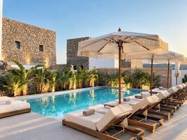 Uma área de piscina com espreguiçadeiras e guarda-sóis, cercada por paredes de pedra e plantas tropicais.