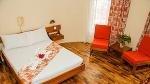 Quarto de hotel com cama de madeira, mesa de cabeceira, cadeira vermelha e pequena mesa com uma planta.