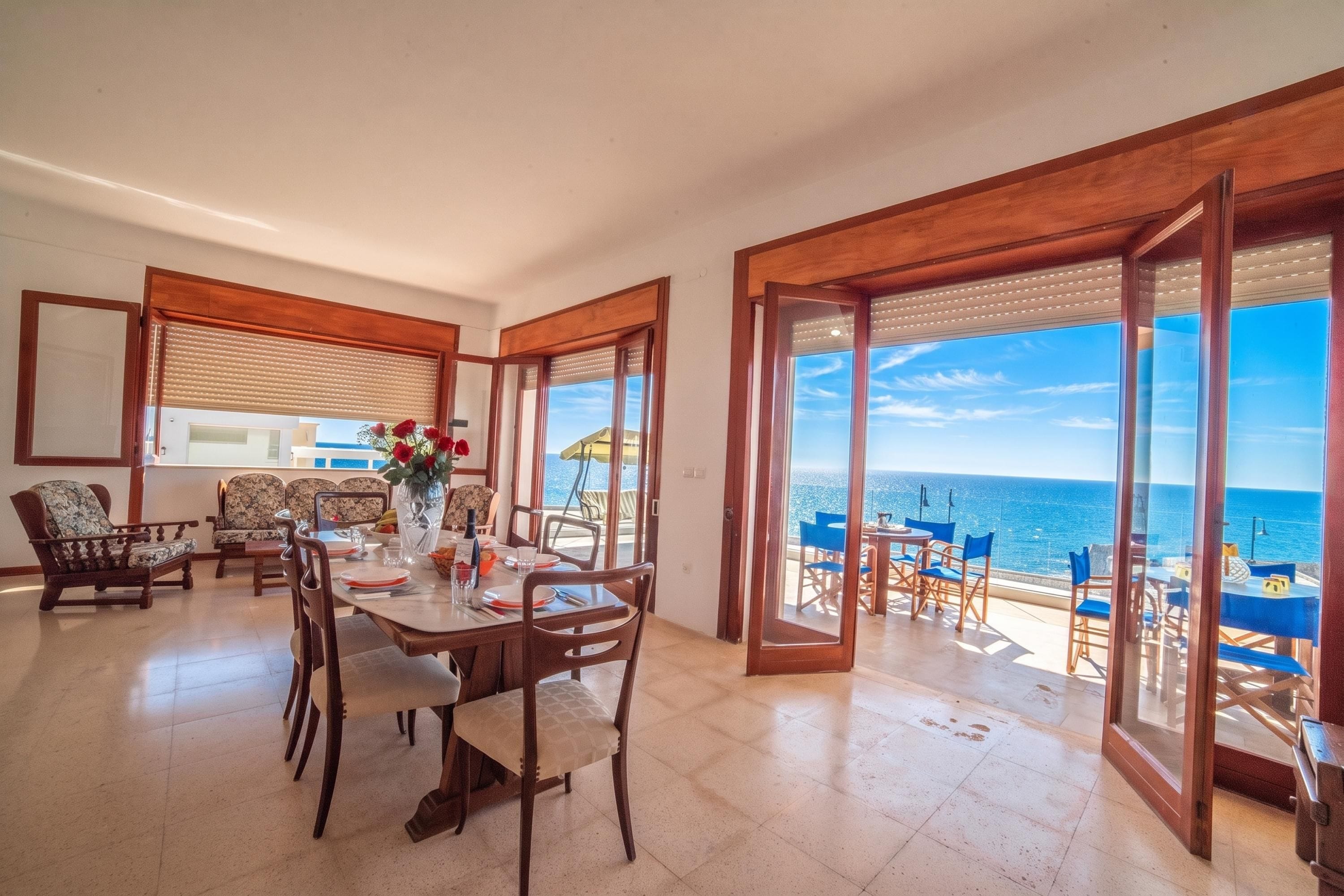 Uma área de refeições com uma mesa posta para quatro pessoas, um vaso com flores e vista para o mar através de portas abertas.