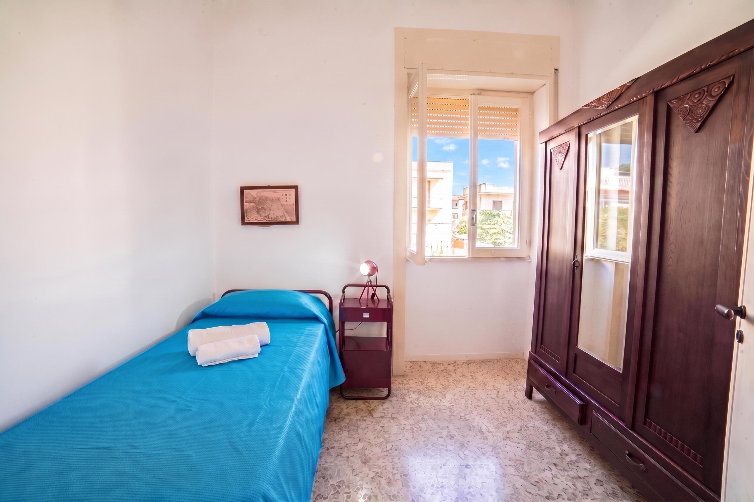 Um quarto pequeno com cama, guarda-roupa, mesa de cabeceira e janela.