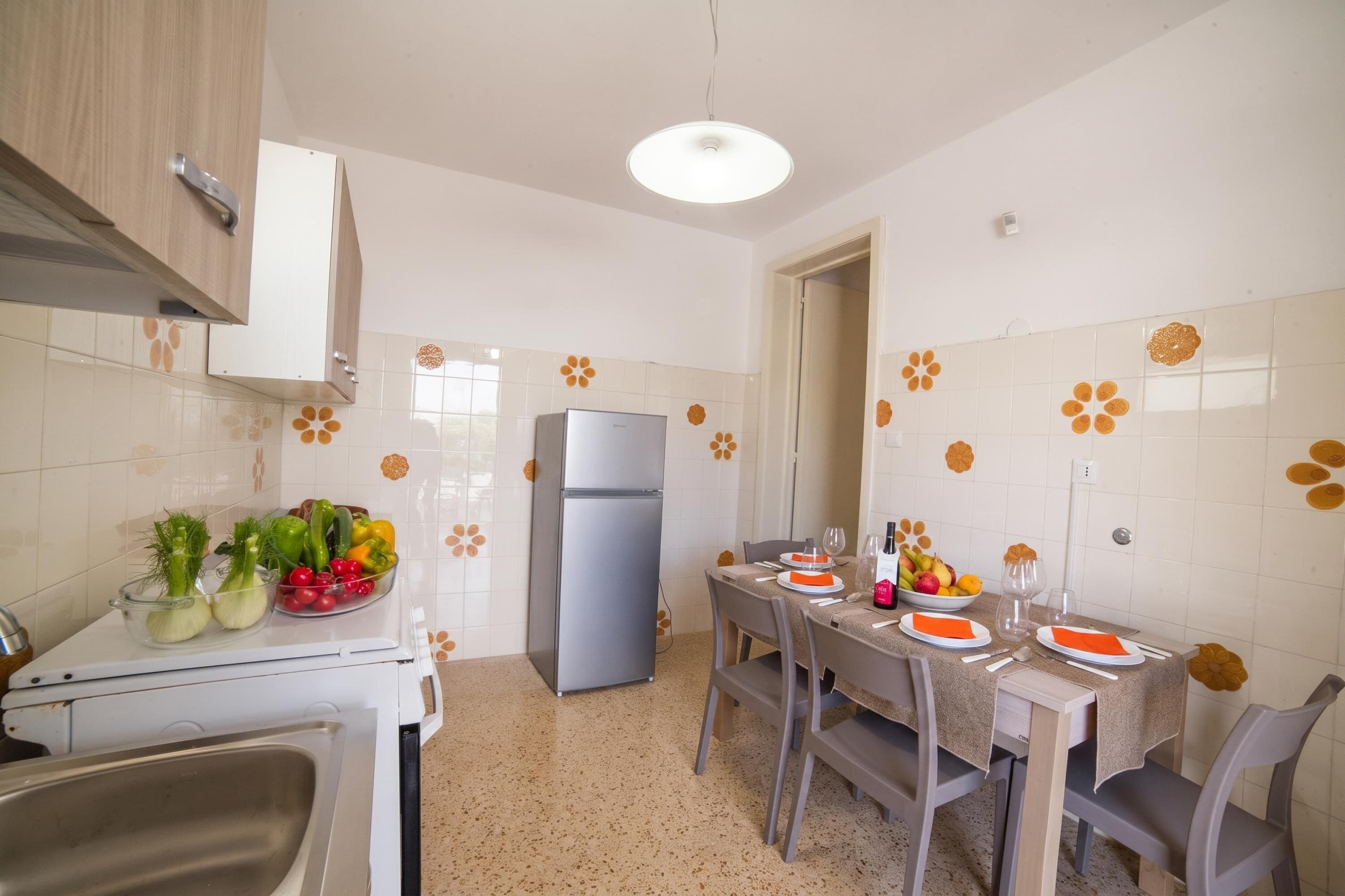 Uma cozinha moderna com área de refeições, equipada com geladeira, pia e mesa posta para dois, incluindo pratos, talheres e uma tigela com frutas.