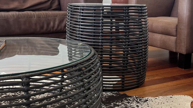 Uma sala de estar com um sofá de couro marrom, duas mesas de centro de vime, uma mesa com tampo de vidro, uma planta em vaso e um tapete de couro.