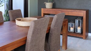 Uma sala de jantar com mesa de madeira, cadeiras estofadas cinzas, luminárias pendentes, um espelho grande e um vaso de planta.