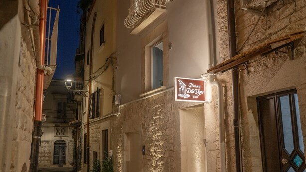 Uma rua estreita com calçada de pedra, um edifício com a placa "Olimpia 101 Della Lira" e uma escadaria que leva a uma entrada.
