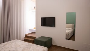 Quarto com cama, um banquinho verde, televisão na parede e um espelho refletindo a cama.