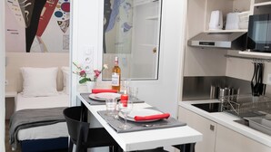 Quarto de hotel moderno com cama, uma mesa de jantar posta para dois, cozinha compacta com micro-ondas e um espelho grande que reflete o ambiente.