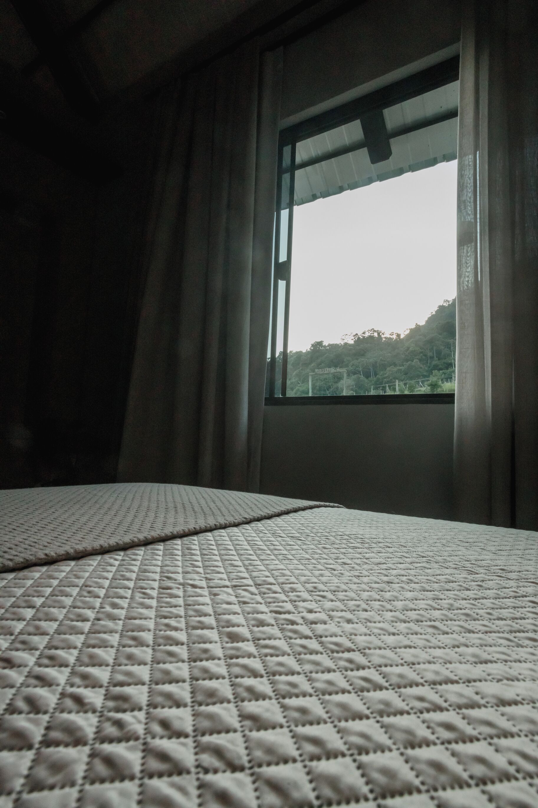 Uma cama com manta acolchoada, uma janela com cortinas e vista para uma paisagem verde.