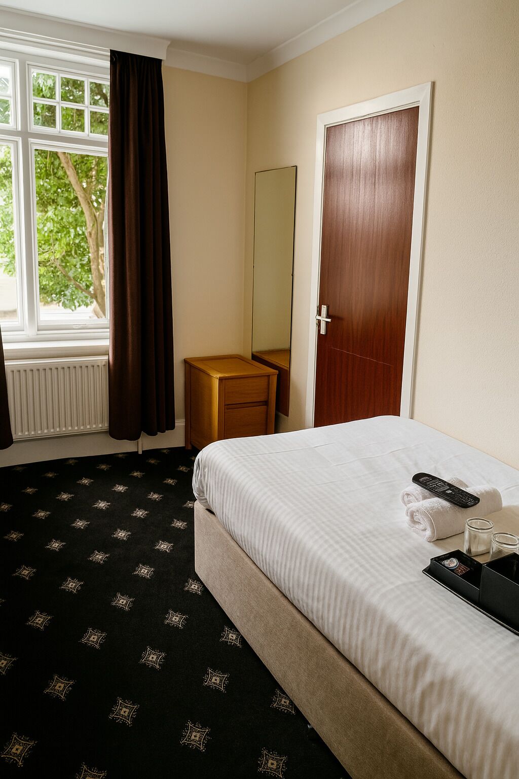 Quarto de hotel com uma cama, uma mesinha de cabeceira de madeira, um espelho, uma porta e uma janela com cortinas.