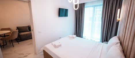 Quarto de hotel com cama, televisão, área de jantar com mesa e cadeiras, e janela com cortinas.