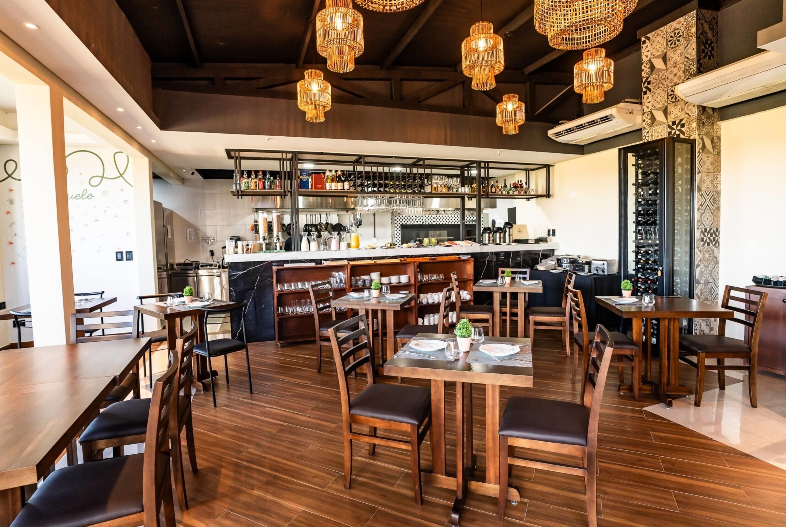 Um restaurante com mesas e cadeiras de madeira, área de bar com prateleiras de garrafas e lustres de vime pendentes.