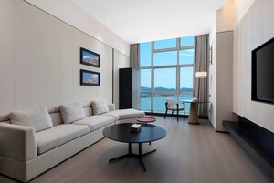 Quarto de hotel moderno com uma janela grande, um sofá, uma mesa de centro, uma TV e uma pequena mesa com abajur.