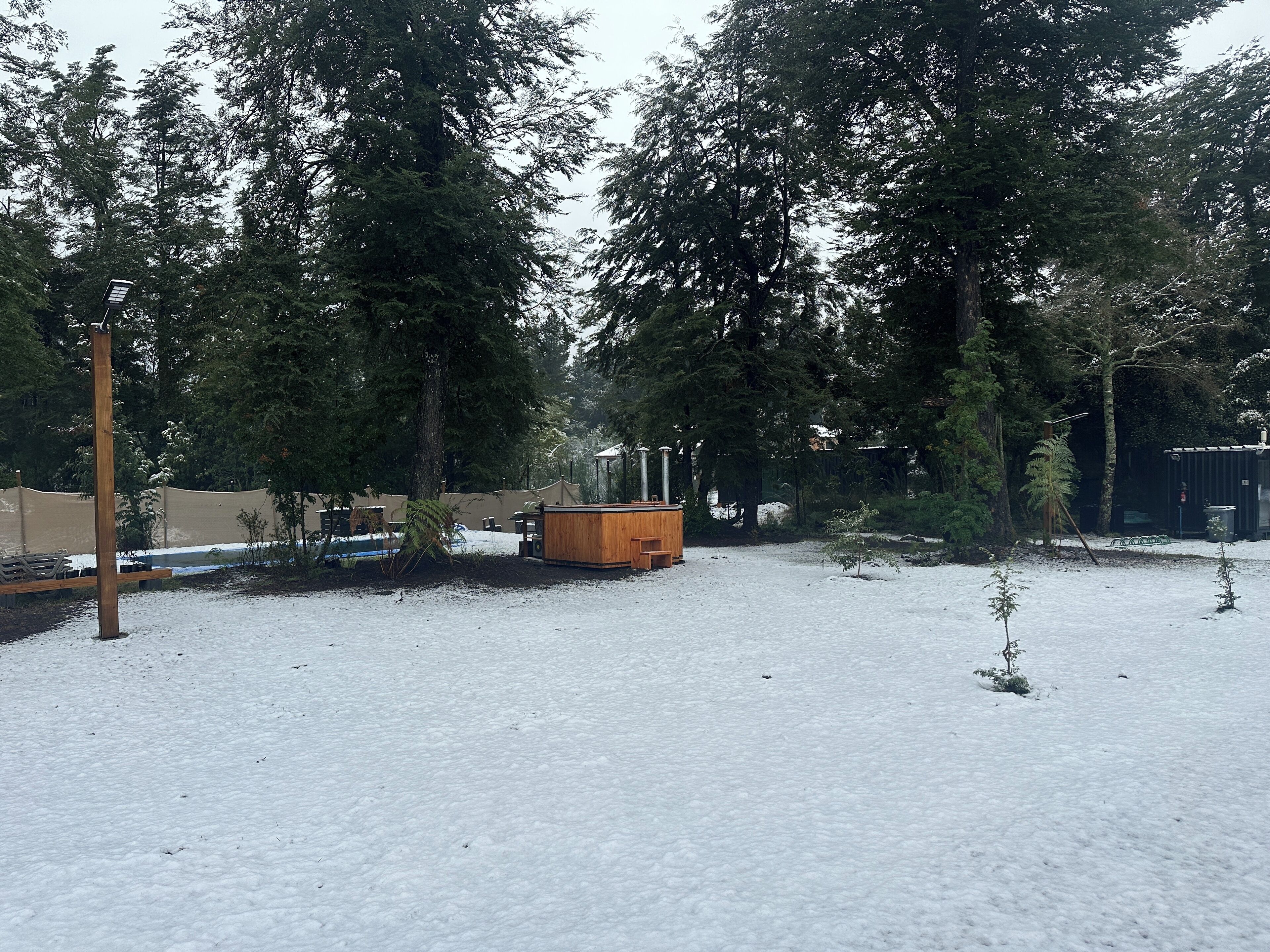 Área externa coberta de neve com uma estrutura de madeira, possivelmente uma sauna ou banheira de hidromassagem, cercada por altas árvores perenes.