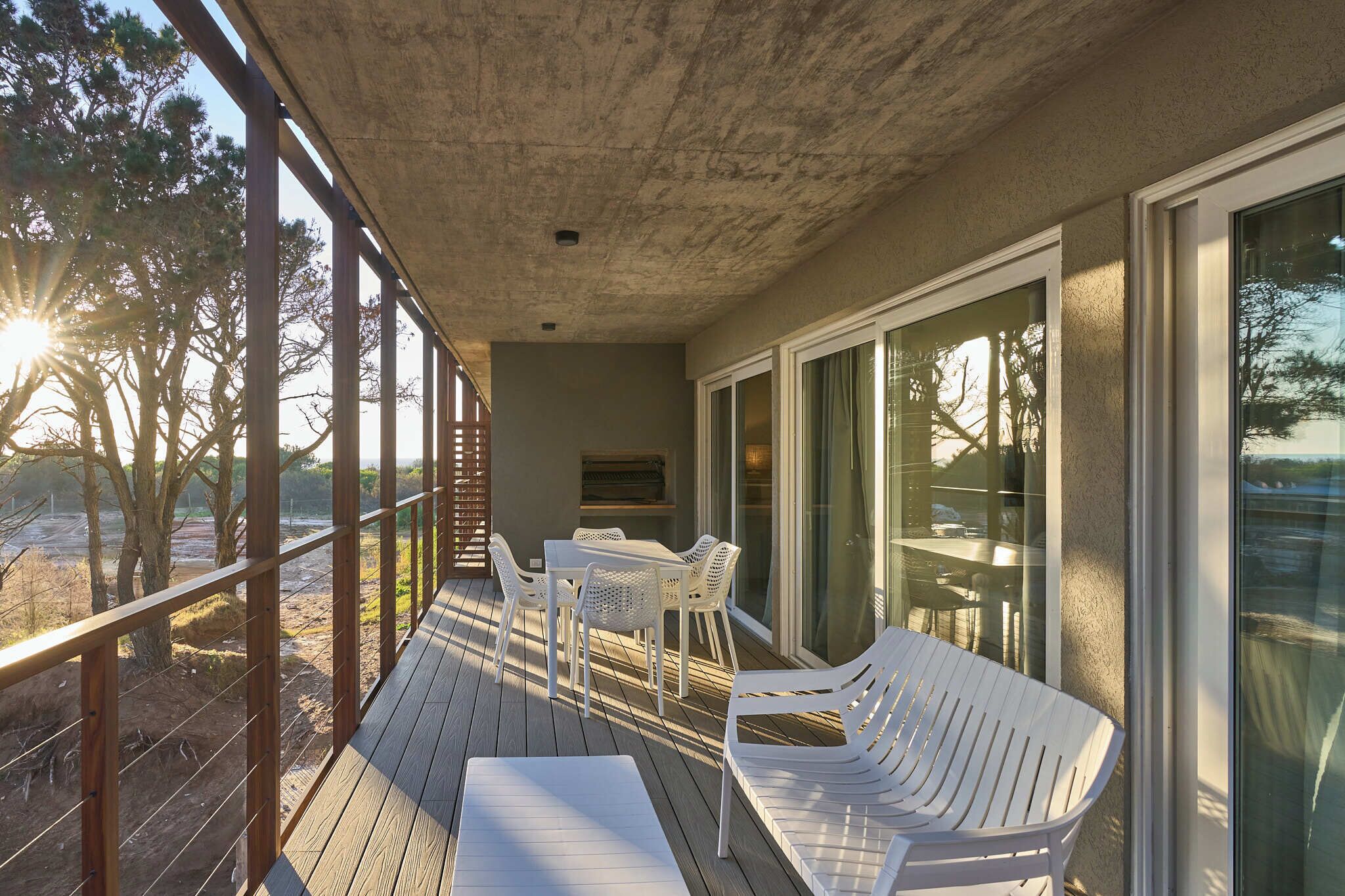 Um terraço coberto ao ar livre com mesa e cadeiras, janelas amplas e vista para árvores e um corpo de água.