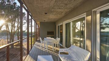 Um terraço coberto ao ar livre com mesa e cadeiras, janelas amplas e vista para árvores e um corpo de água.