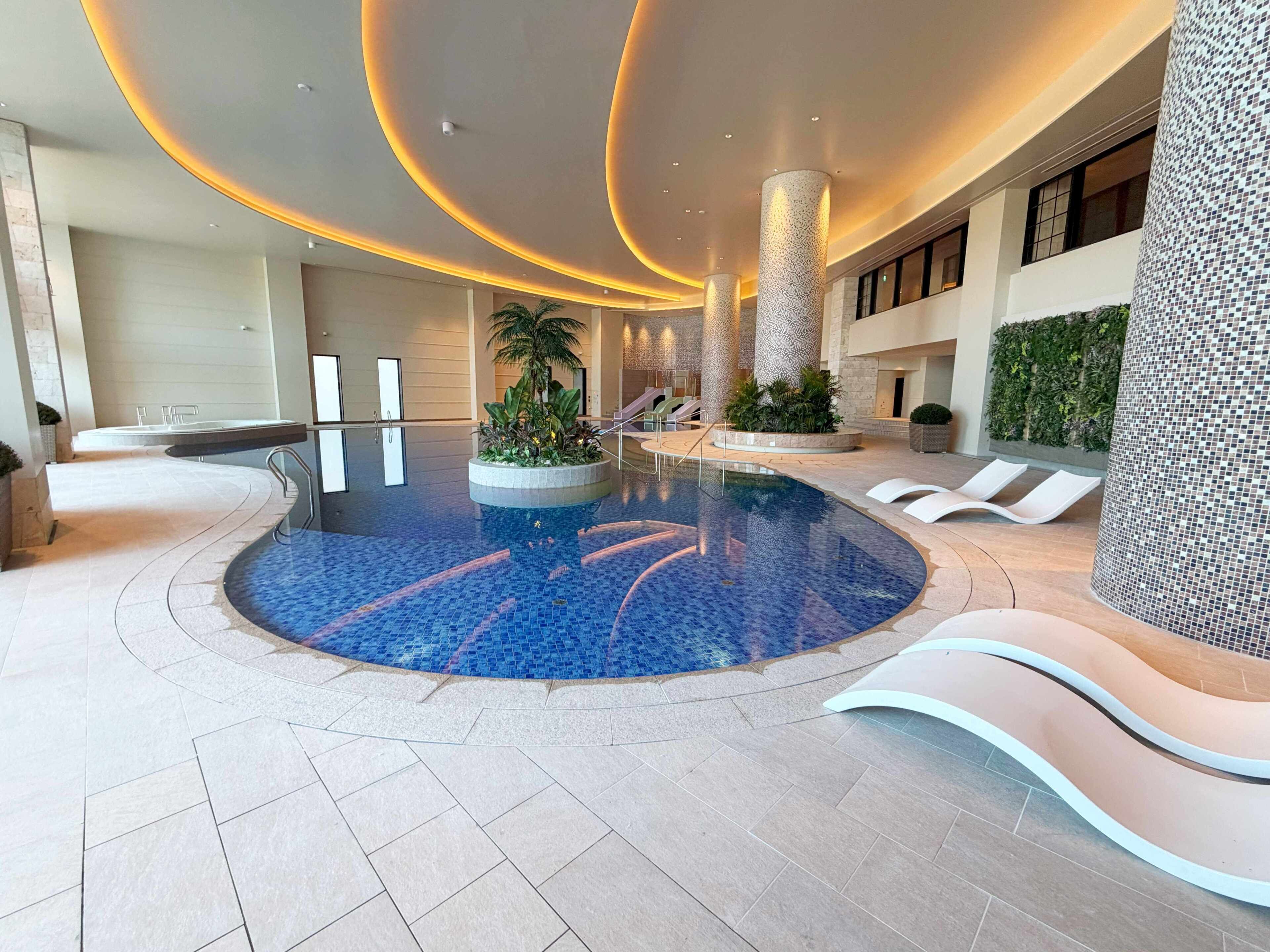 Piscina interior moderna com design curvo, rodeada por espreguiçadeiras e plantas em vasos.