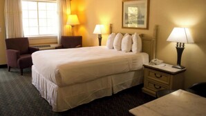 Quarto de hotel com uma cama grande, duas luminárias de cabeceira, uma cadeira, uma escrivaninha e um quadro na parede.