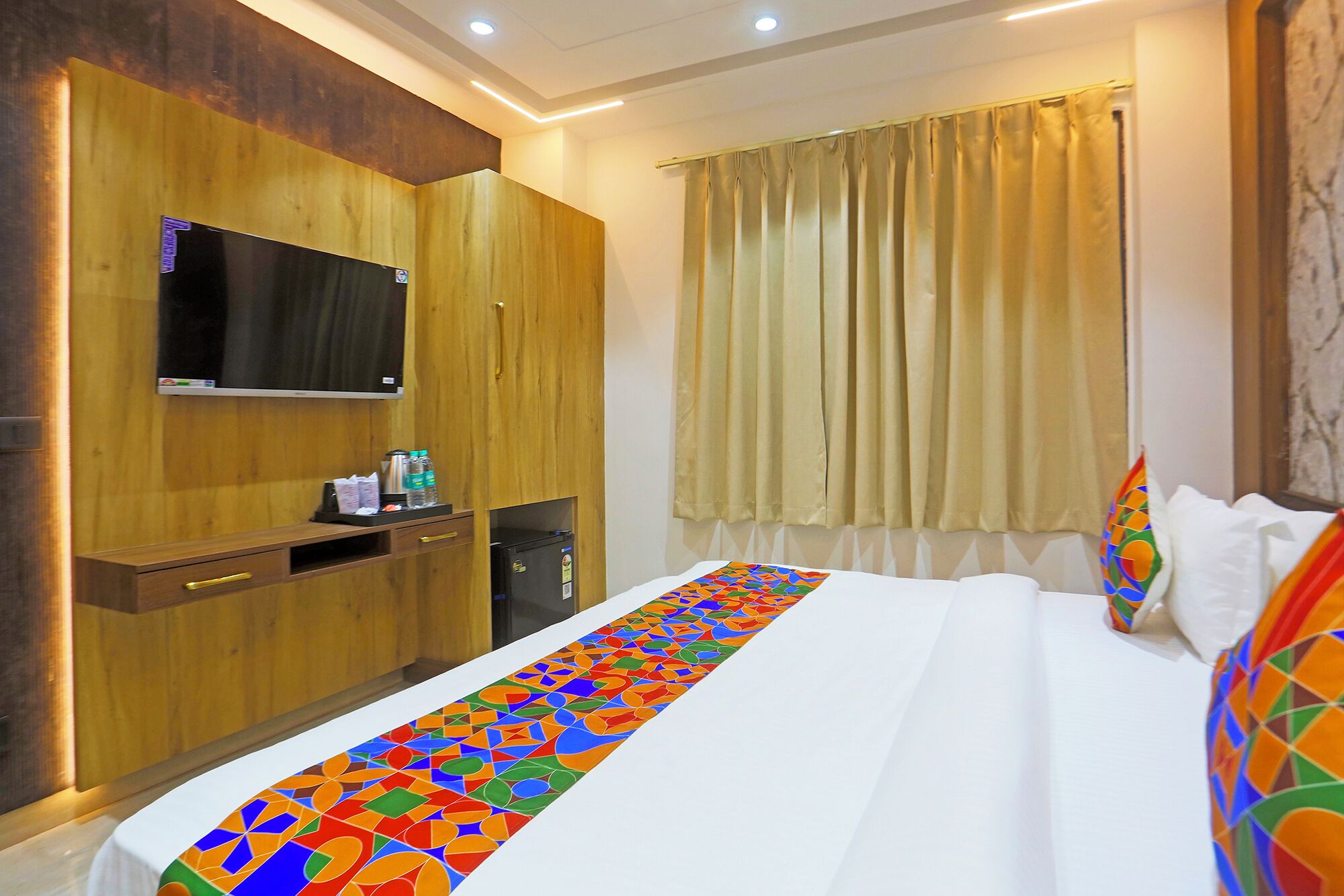 Quarto de hotel com cama, televisão fixada num móvel de madeira, secretária com cadeira e janela com cortinas.