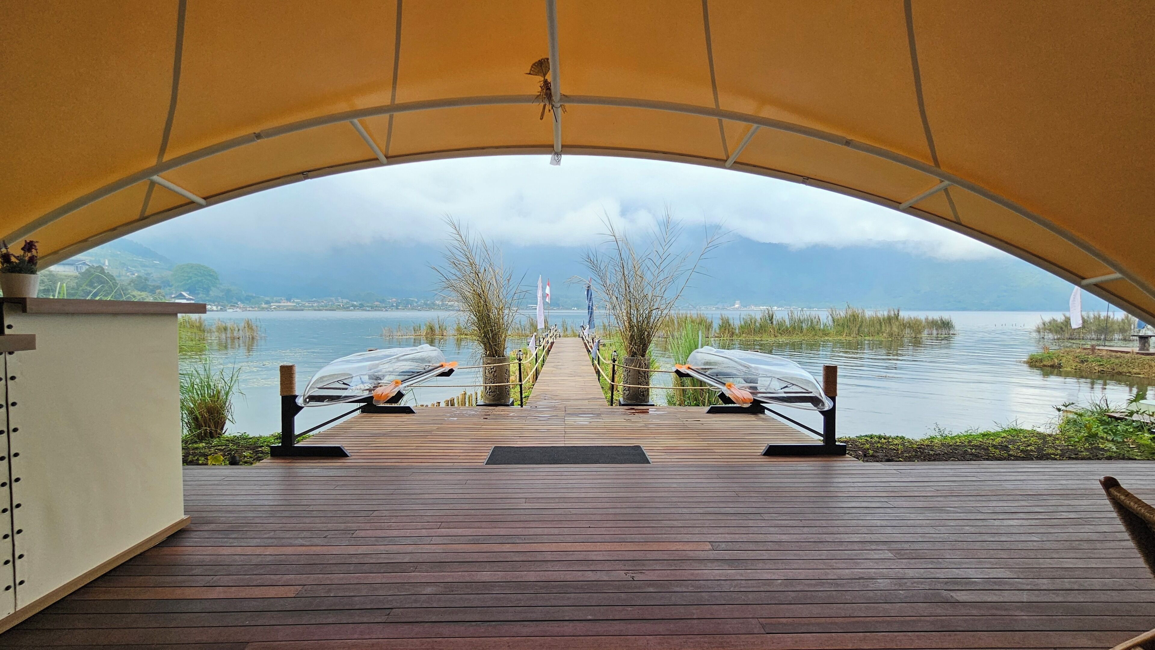 Um deck de madeira com toldo, com vista para um lago calmo e montanhas ao longe.