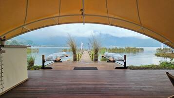 Um deck de madeira com toldo, com vista para um lago calmo e montanhas ao longe.