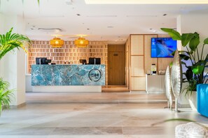 Uma área de recepção de hotel com balcão de mármore, painéis de madeira, televisão e plantas em vasos.