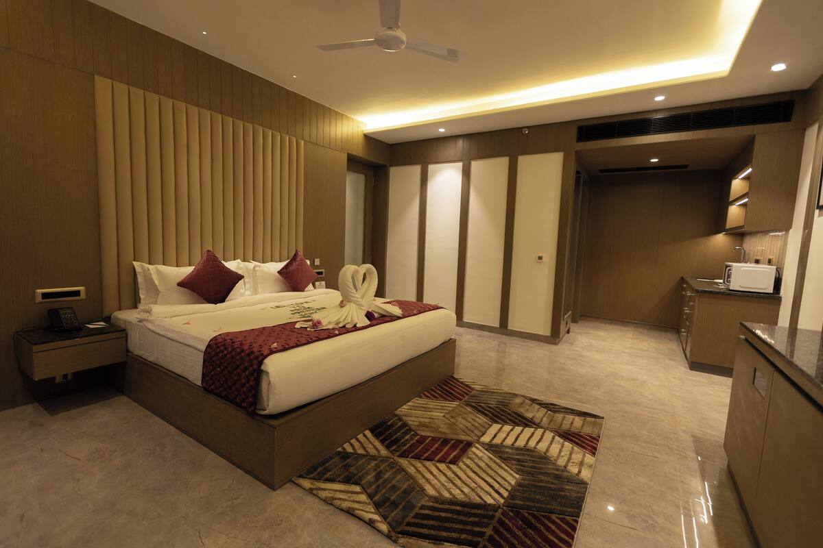 Quarto de hotel moderno com uma cama grande, uma mesa de cabeceira, um abajur, um ventilador de teto, um tapete estampado e um banheiro com pia e espelho.