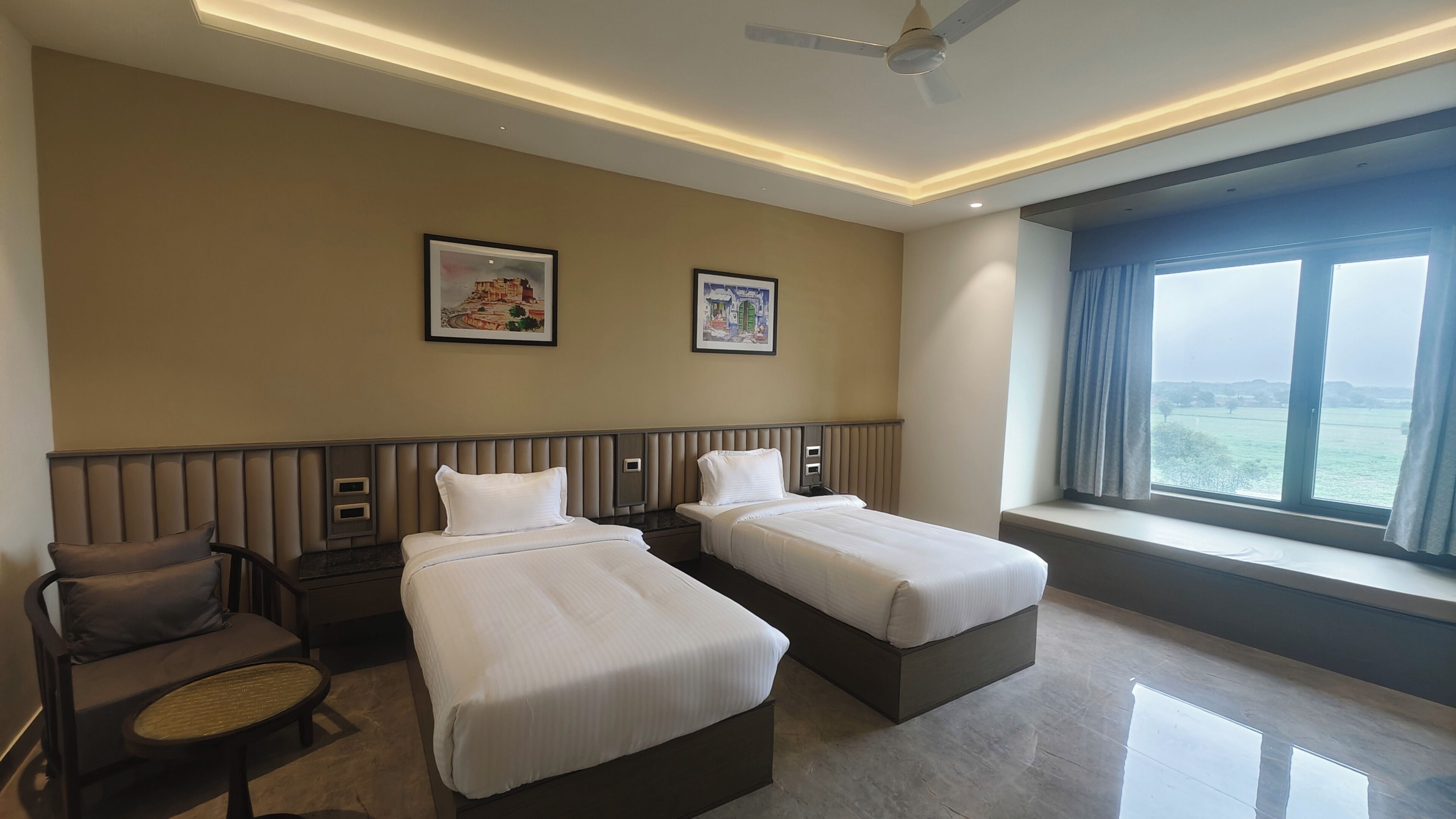 Quarto de hotel com duas camas, uma cadeira, uma mesa pequena, um ventilador de teto e uma janela com vista.