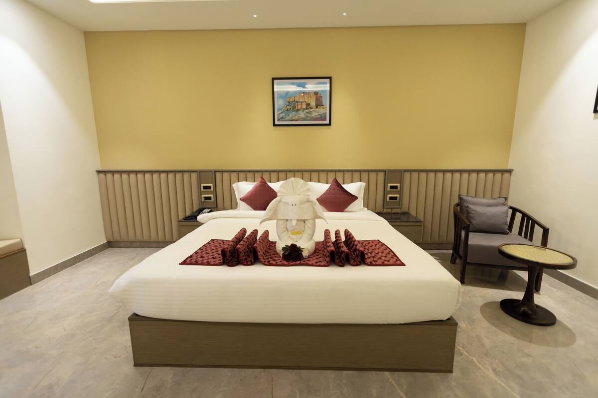 Quarto de hotel com uma cama grande, cabeceira com ripas verticais, mesa de cabeceira com abajur, uma cadeira e uma mesinha redonda.