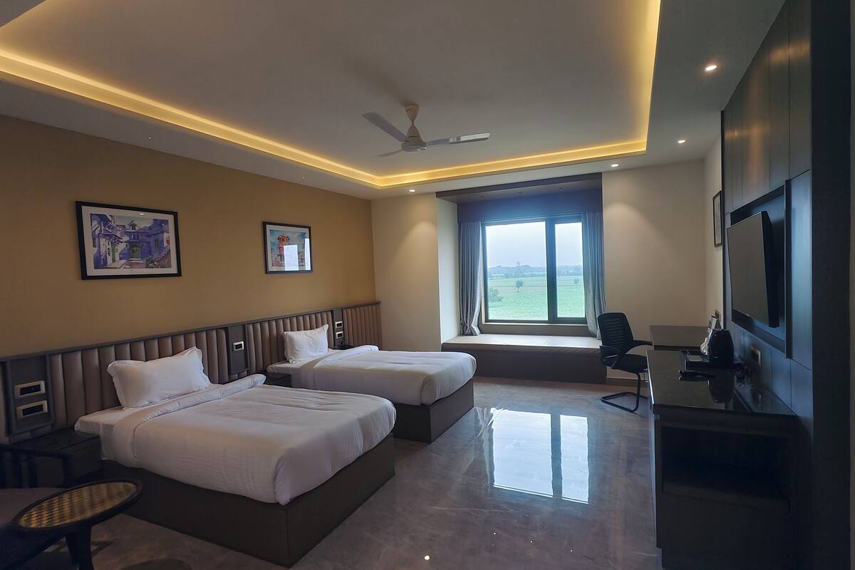 Quarto de hotel com duas camas, uma escrivaninha, uma cadeira, televisão e uma grande janela com vista para o campo.
