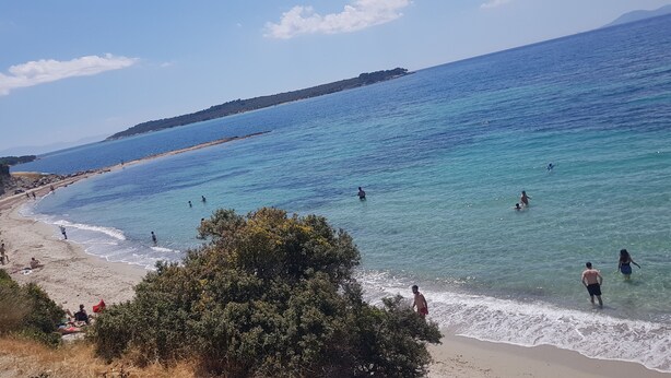 Uma praia com águas azuis cristalinas, pessoas nadando e caminhando pela orla, e uma massa de terra ao longe.